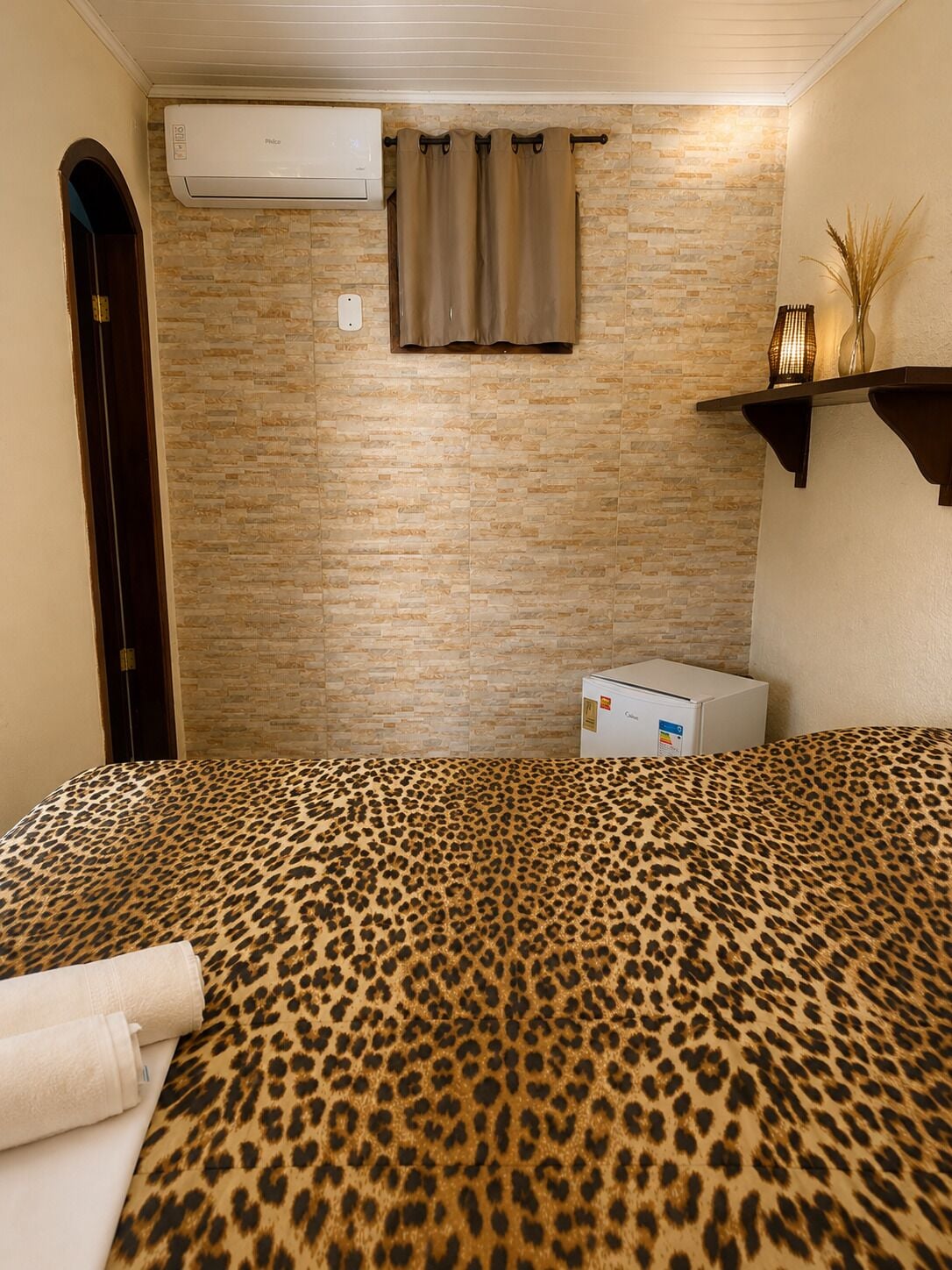 Um quarto com parede de tijolos, uma cama com colcha estampada de leopardo, um ar condicionado, um eletrodoméstico pequeno branco e uma estante com objetos decorativos.
