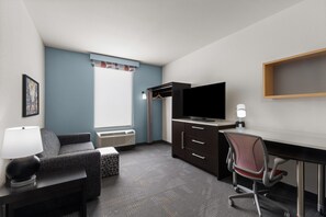 Quarto de hotel com cama, escrivaninha, cadeira, televisão, luminária e janela com cortinas.