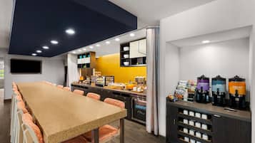 Um café moderno com uma mesa comprida, cadeiras, um balcão com máquinas de café e uma geladeira.
