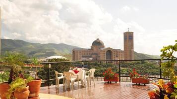Terraço com mesa posta para dois, com vista para um edifício amplo com cúpula e torre do relógio, cercado por vegetação e colinas.