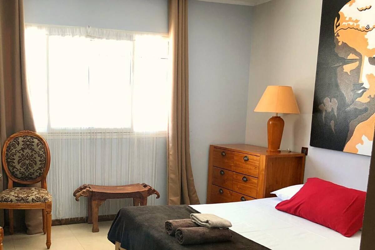 Quarto de hotel com cama, cadeira, mesa de cabeceira de madeira, mesa e janela com cortinas.