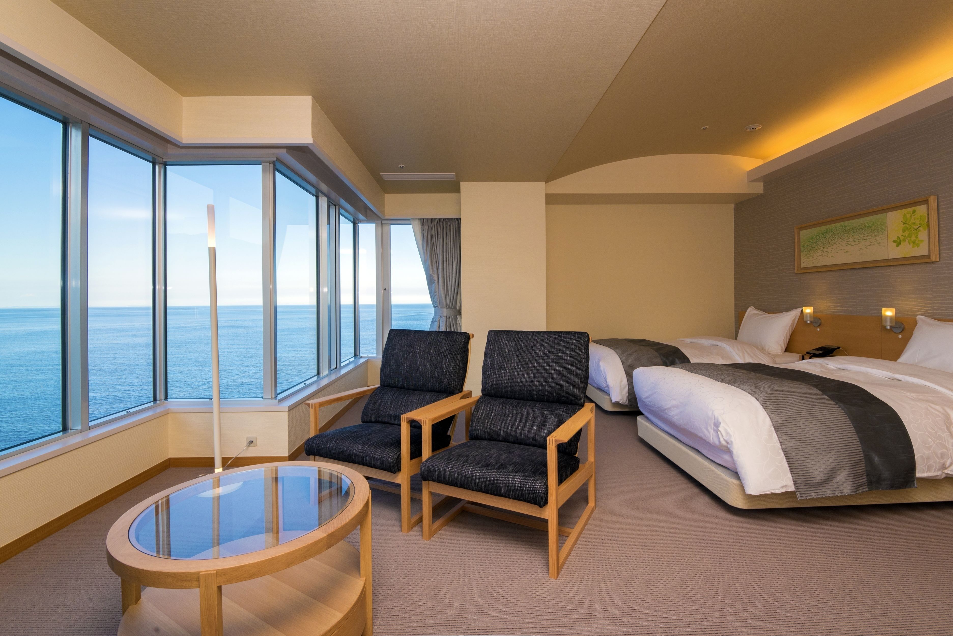 Quarto de hotel com uma janela ampla que oferece vista para o mar, cama com roupa de cama branca, duas poltronas e uma mesa redonda pequena.
