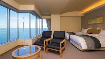 Quarto de hotel com uma janela ampla que oferece vista para o mar, cama com roupa de cama branca, duas poltronas e uma mesa redonda pequena.