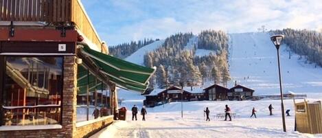 Estância de esqui com um edifício de fachada em pedra e grandes janelas, área de estar coberta ao ar livre e paisagem nevada com pistas de esqui ao fundo.