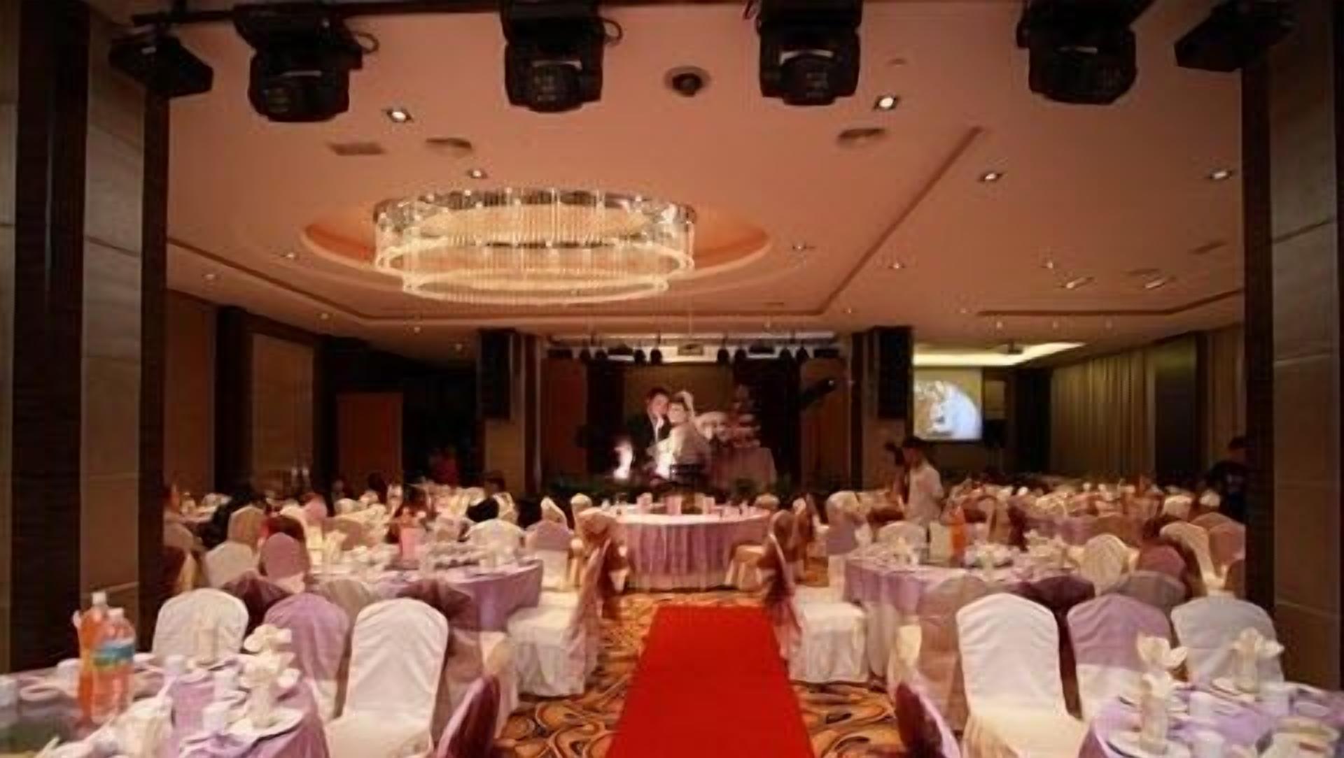 Uma sala de banquetes com mesas redondas cobertas por toalhas brancas e cadeiras com capas brancas. Um tapete vermelho percorre o centro do espaço.