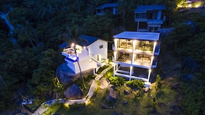 Uma casa moderna de dois andares com telhado plano, rodeada por vegetação exuberante e pedras.
