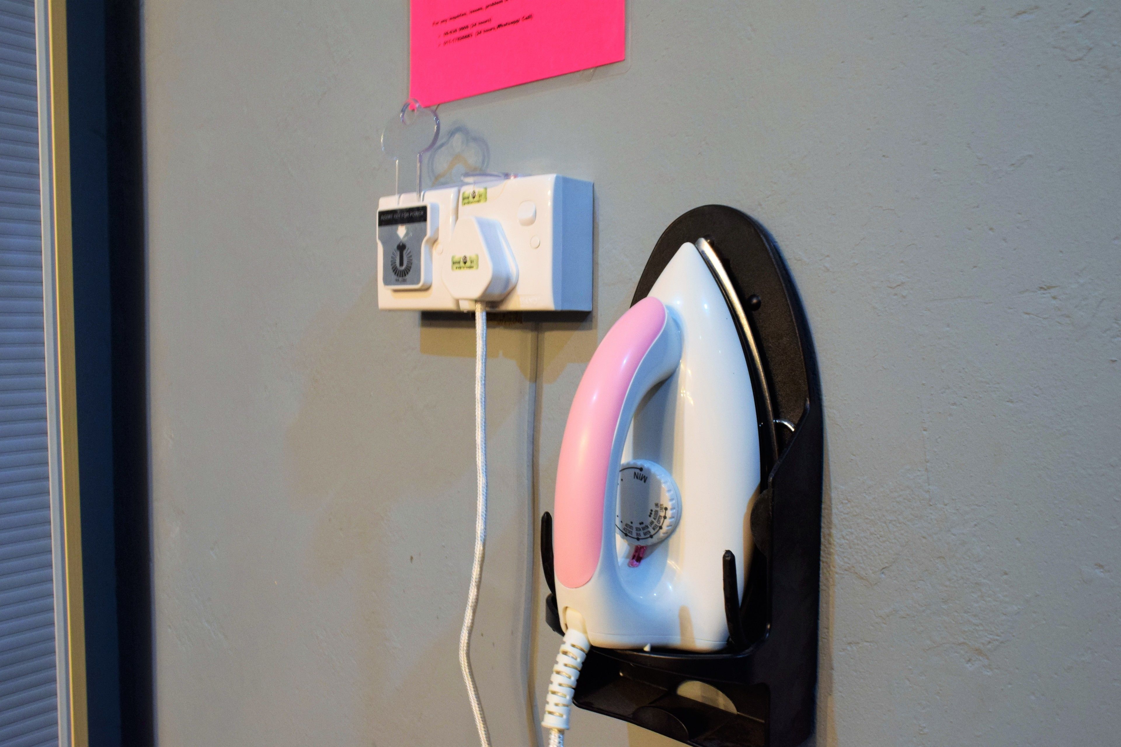 Um telefone fixo com fio, um pequeno aparelho com tela e um bilhete rosa na parede.