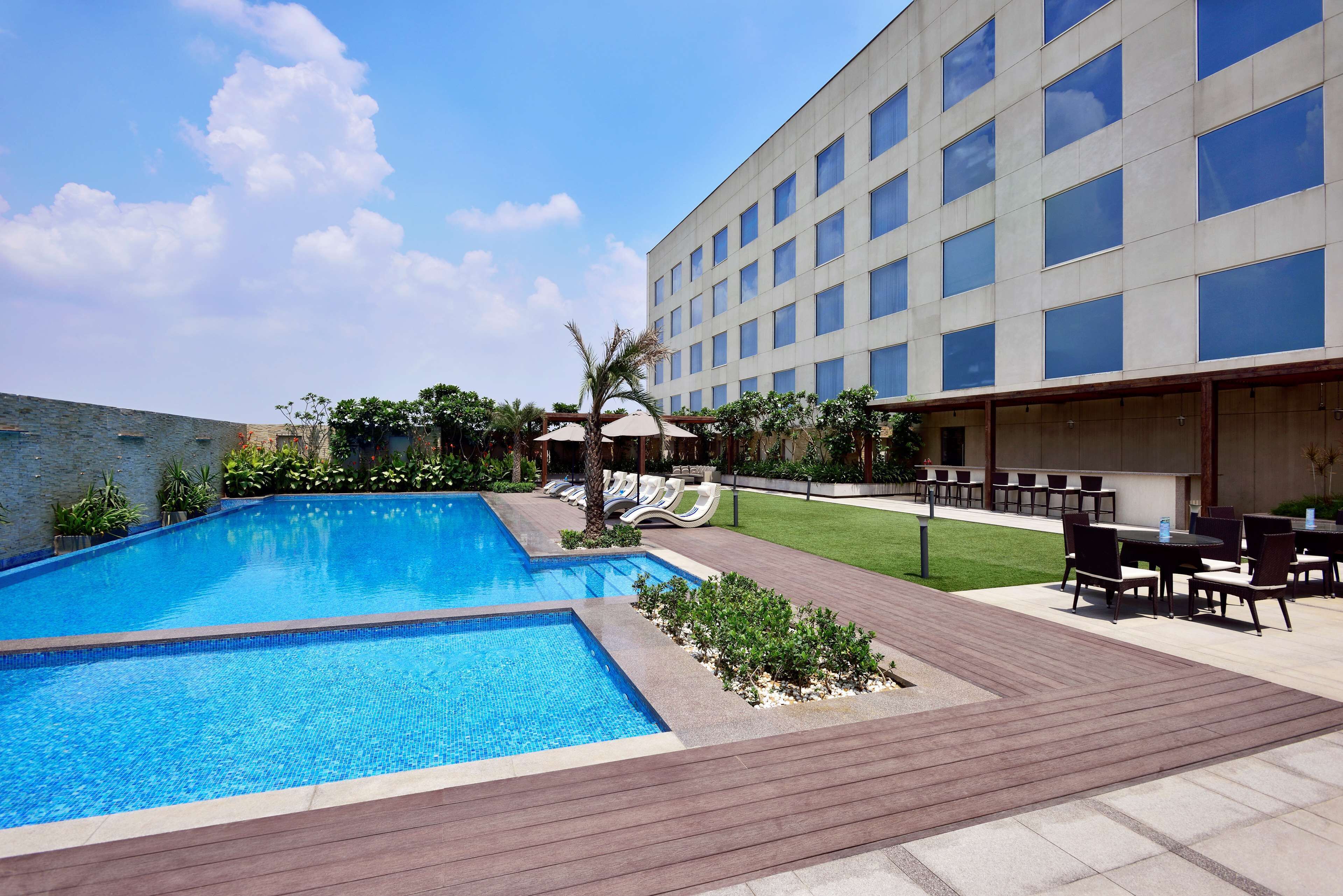 Piscina com deck de madeira, área de estar externa e um edifício com grandes janelas.