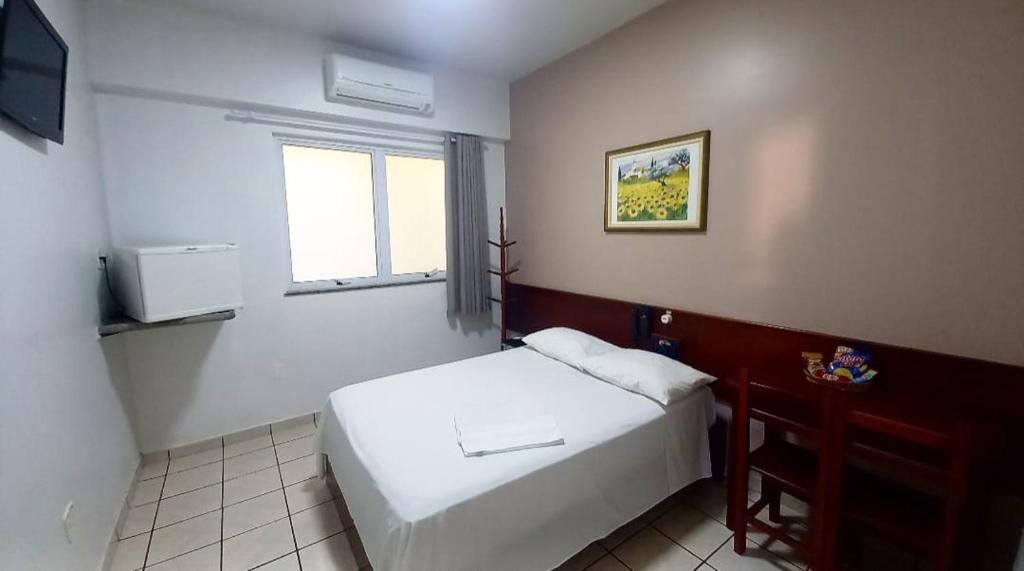 Quarto de hotel com cama, janela, ar condicionado fixado na parede, mesa pequena e uma cadeira.