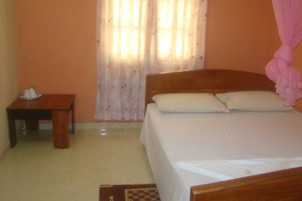 Quarto com cama de madeira, mesa de cabeceira, mesa e janela com cortinas.