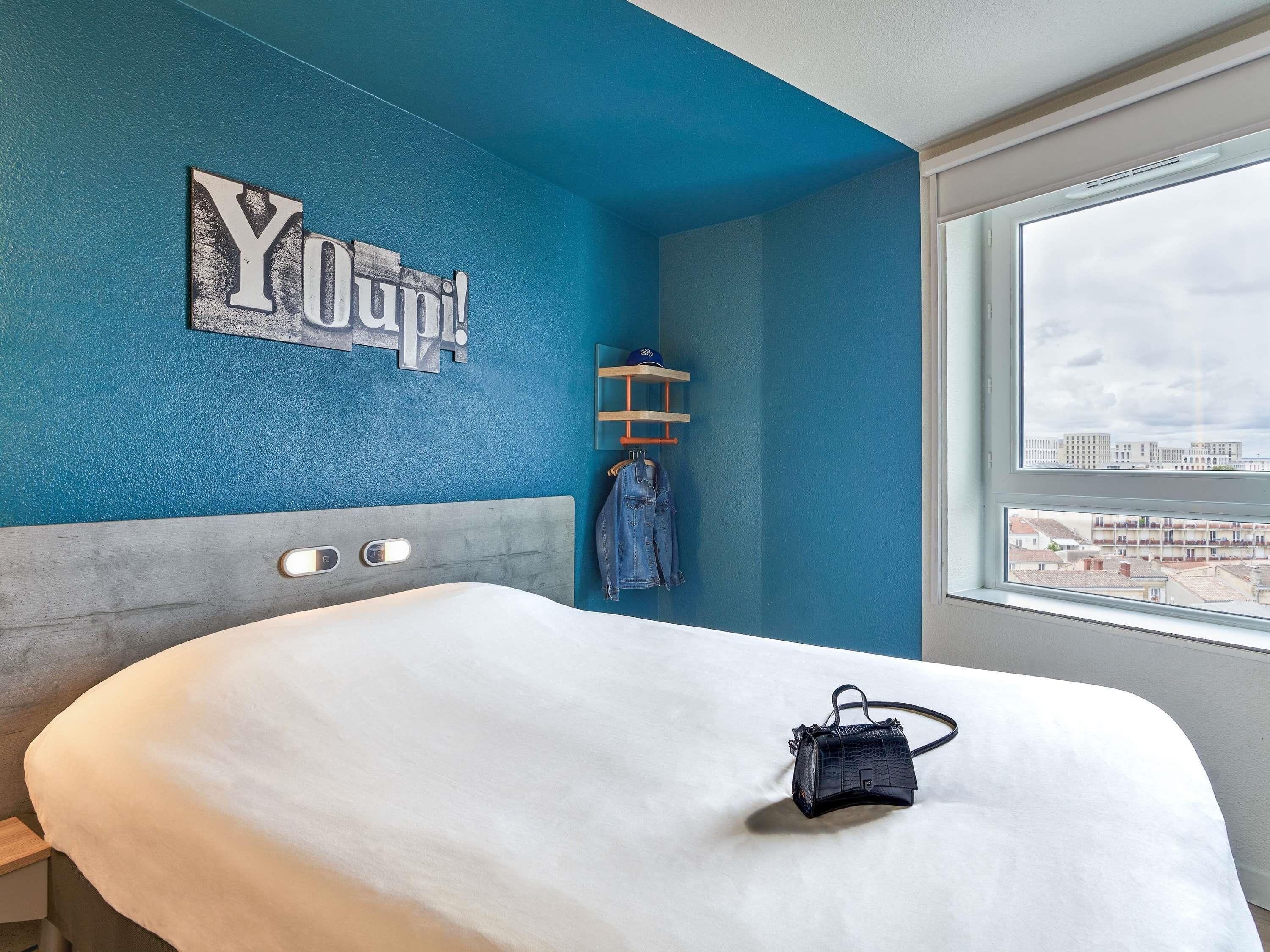 Quarto de hotel com uma cama, um letreiro com a palavra 'Você!', vista de edifícios pela janela e um cabideiro.