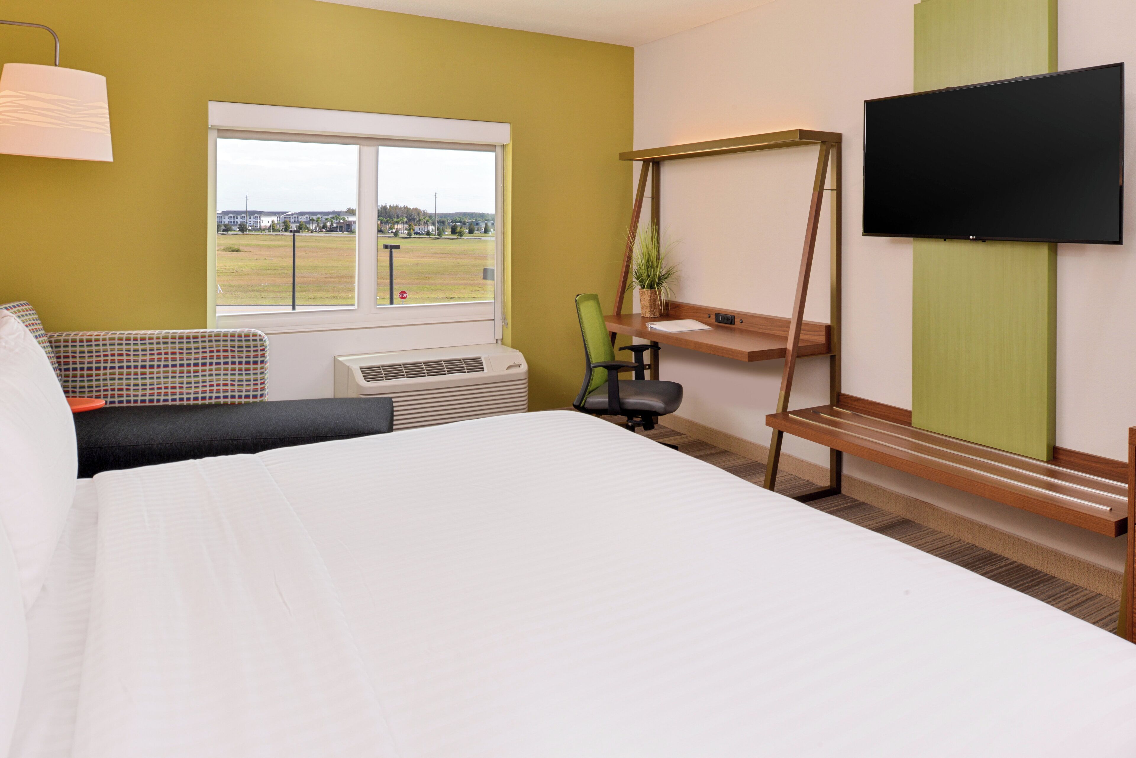 Quarto de hotel com cama, escrivaninha, cadeira, televisão e janela com vista para um campo.