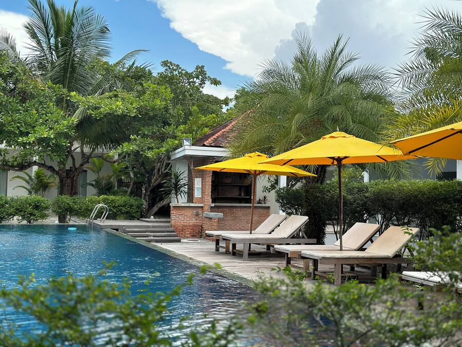 Uma área de piscina com espreguiçadeiras e guarda-sóis amarelos, cercada por vegetação exuberante e um céu claro.