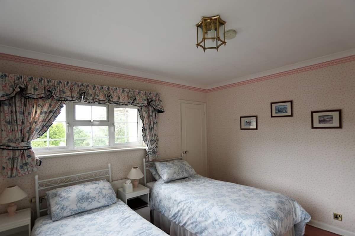Quarto com duas camas de solteiro, janela com cortinas florais, mesa de cabeceira com abajur e luminária na parede.
