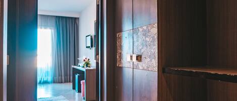 Uma porta de madeira que dá para um quarto com piso ladrilhado, um tapete, uma escrivaninha com um vaso de flores e uma janela com cortinas.