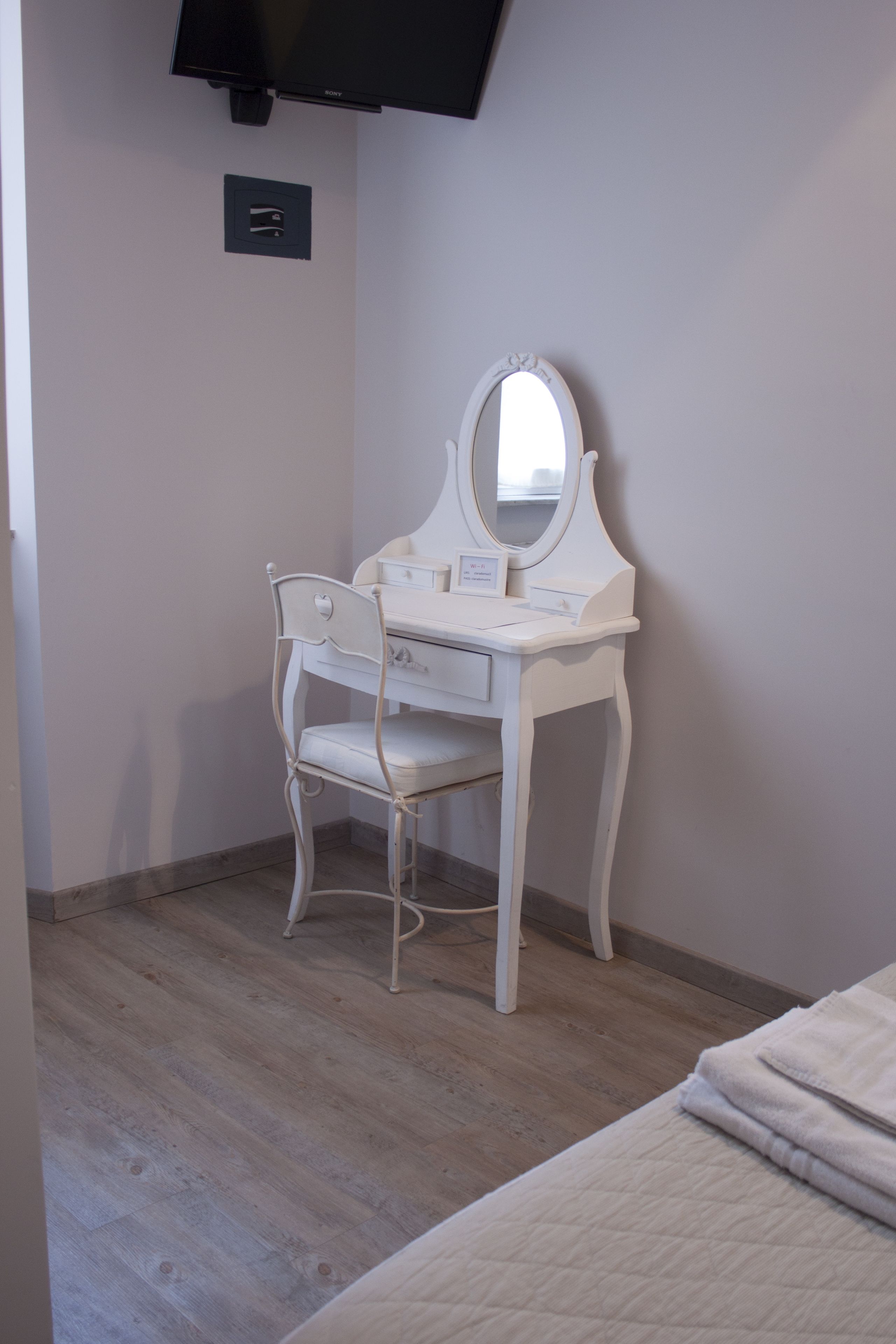 Uma toucador branca com espelho e cadeira, uma TV de tela plana na parede, e uma cama com cobertor branco.
