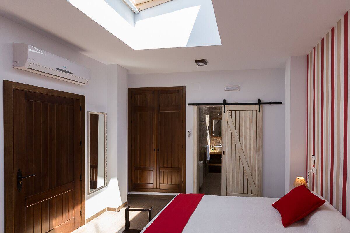 Quarto com claraboia, guarda-roupa de madeira e cama com travesseiro vermelho.