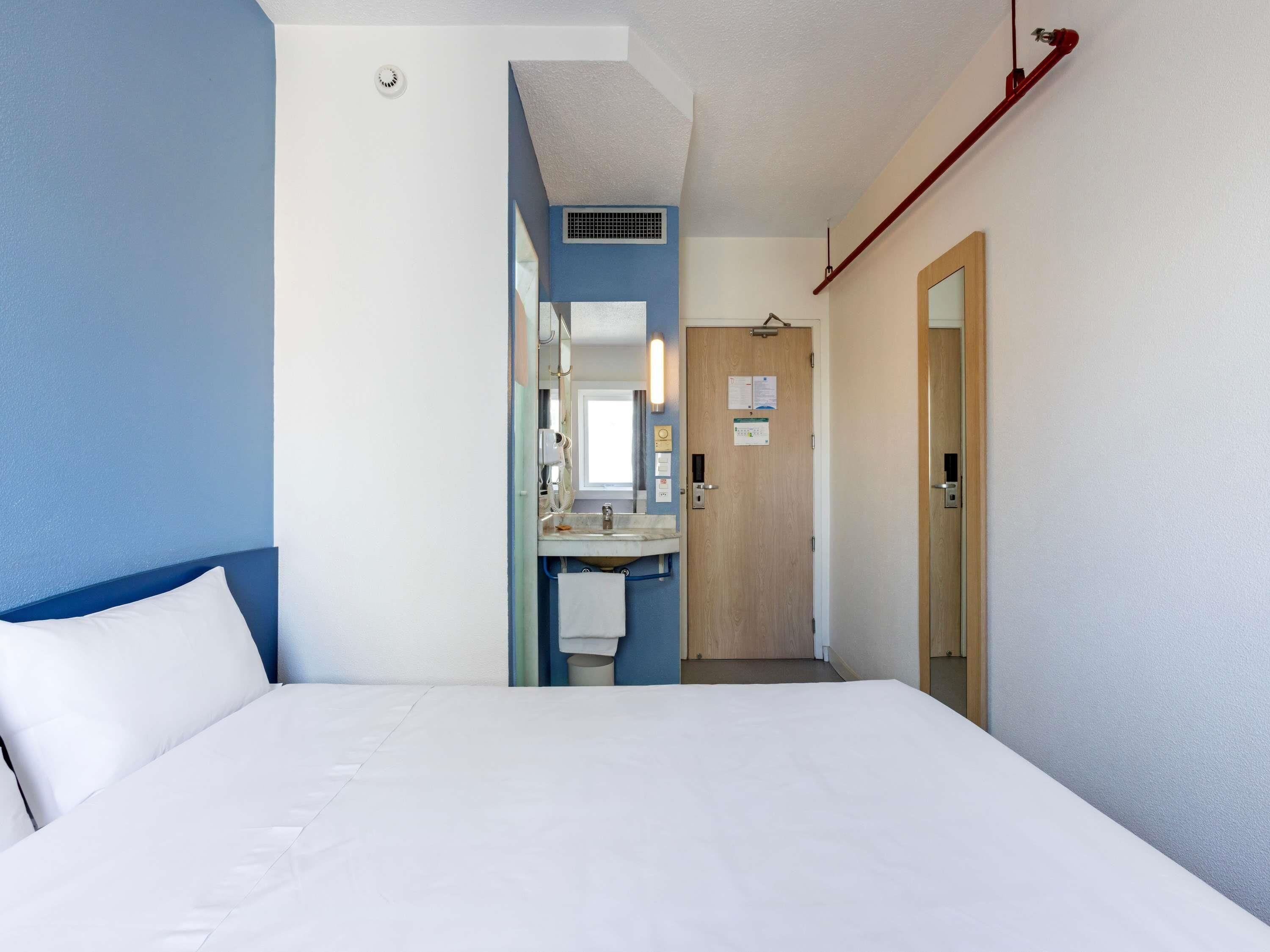 Quarto de hotel com cama, banheiro com pia e espelho, e porta que dá para outro cômodo.