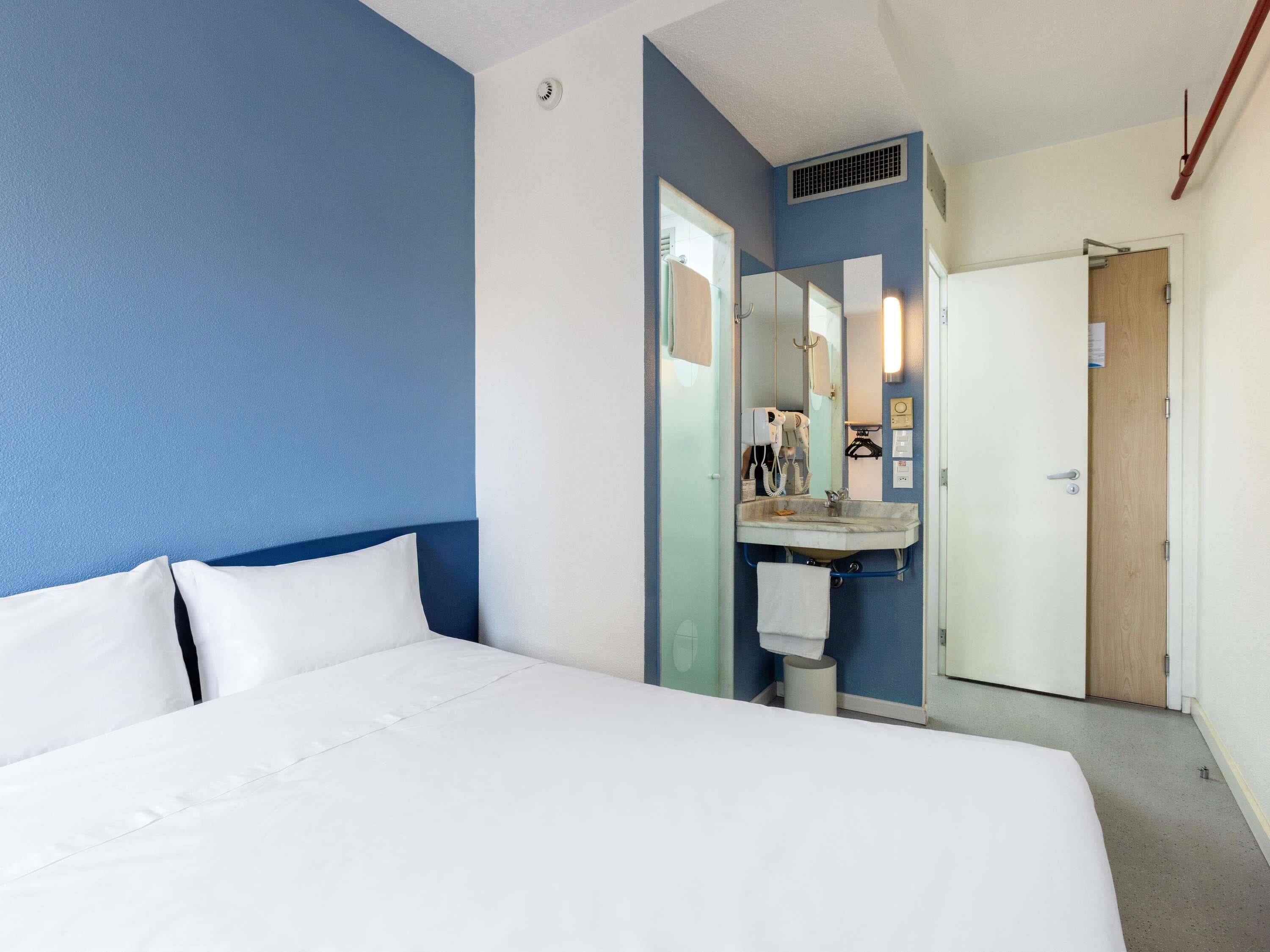 Quarto de hotel com cama, banheiro com pia e espelho, e uma porta.