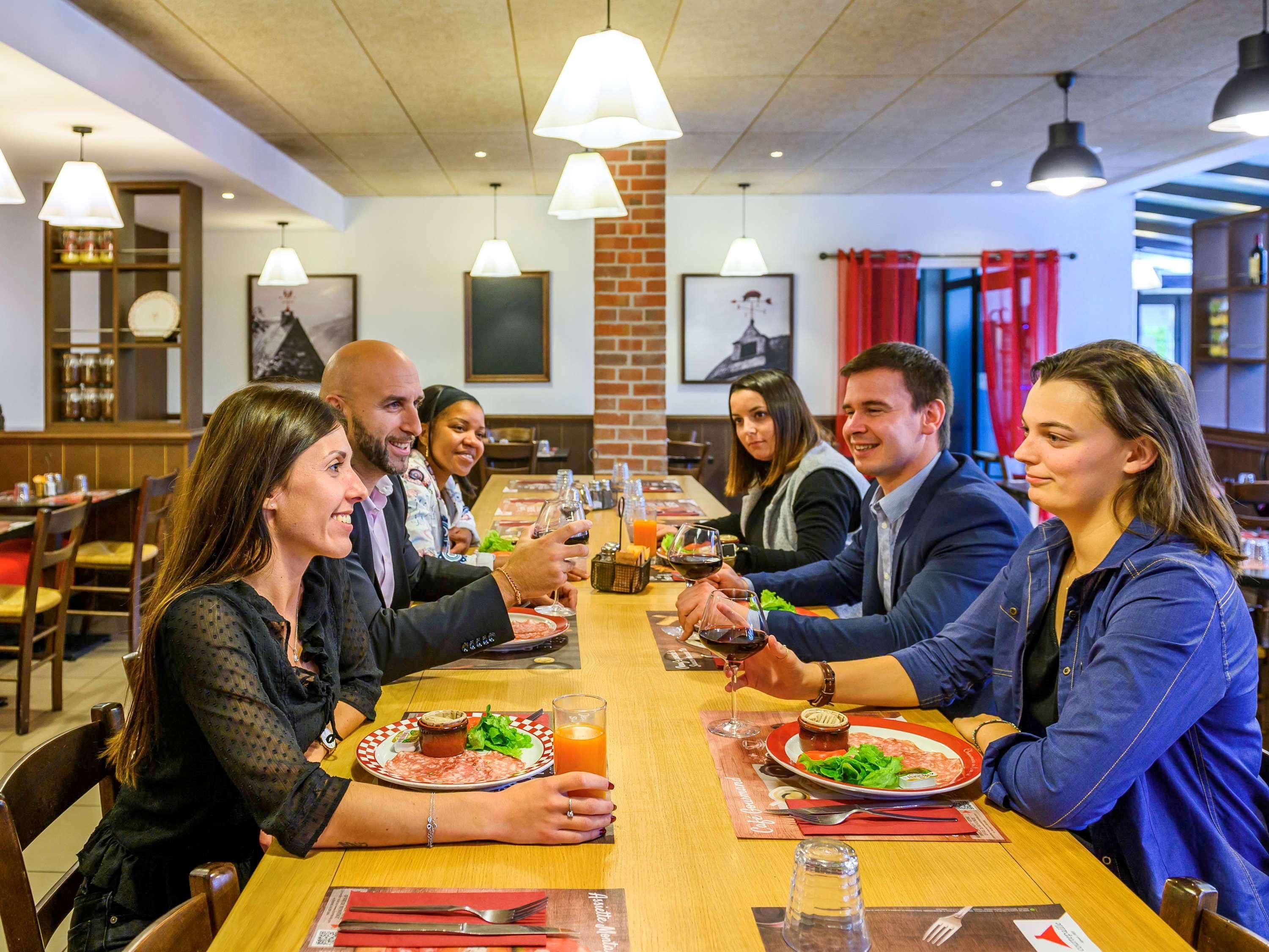 Um grupo de pessoas jantando em um restaurante com uma longa mesa de madeira, luminárias pendentes brancas e uma parede de tijolos ao fundo.