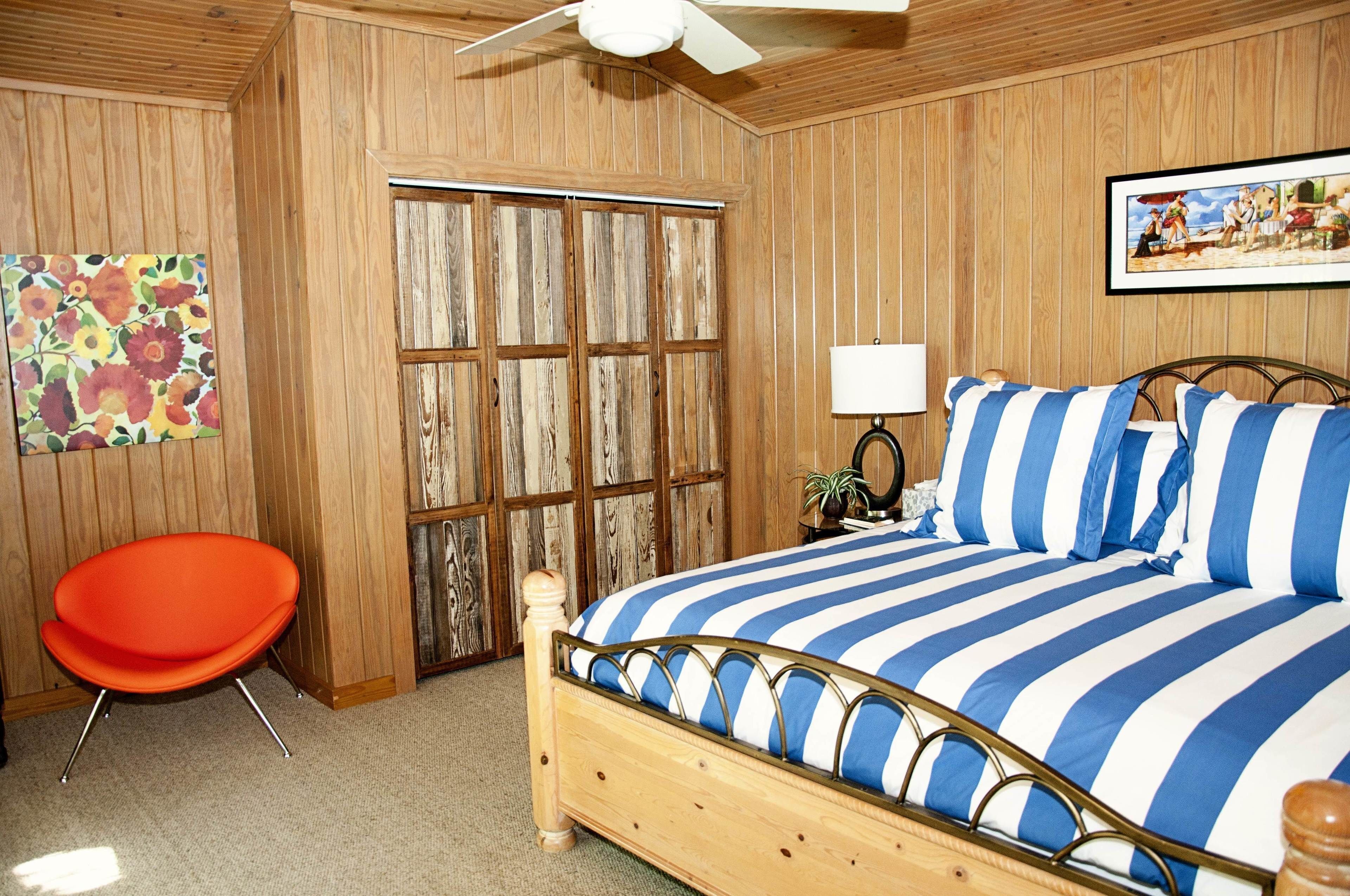 Quarto com cama de madeira, uma cadeira vermelha, ventilador de teto, janela e um quadro na parede.