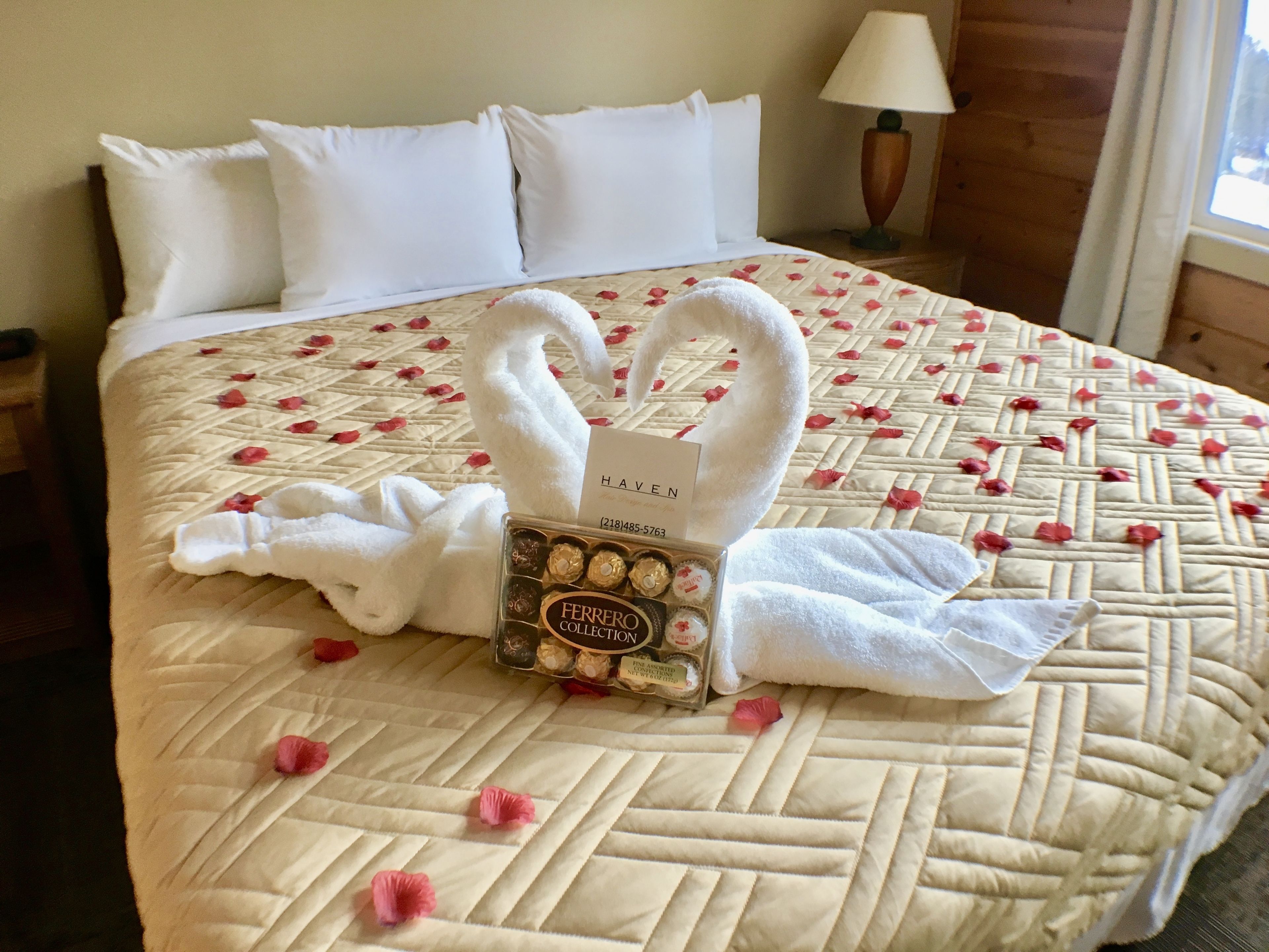Quarto de hotel com uma cama, almofadas brancas, abajur e um arranjo de pétalas de rosa sobre a cama.