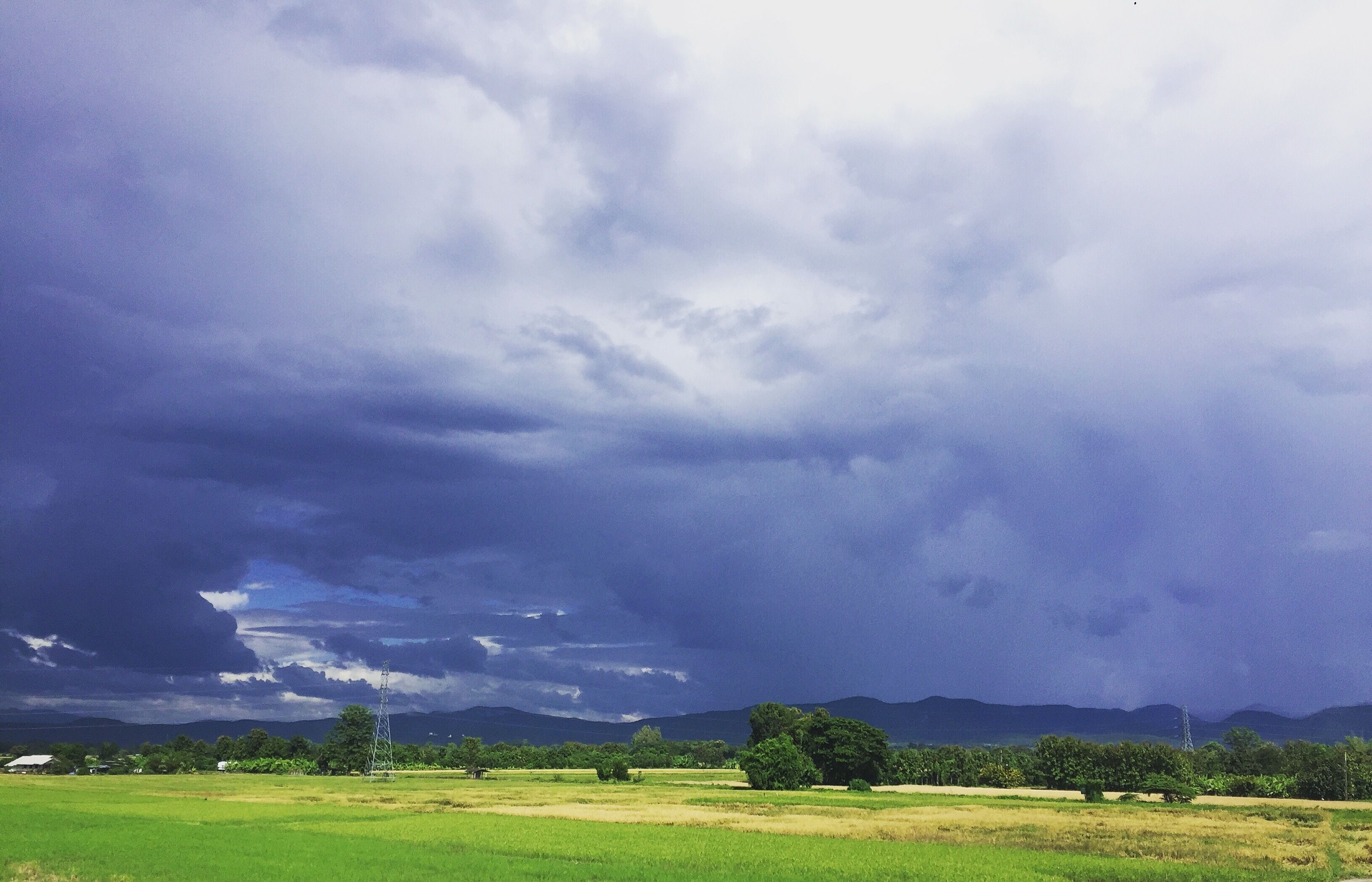 Paisagem rural com campos, árvores e montanhas distantes sob um céu nublado.