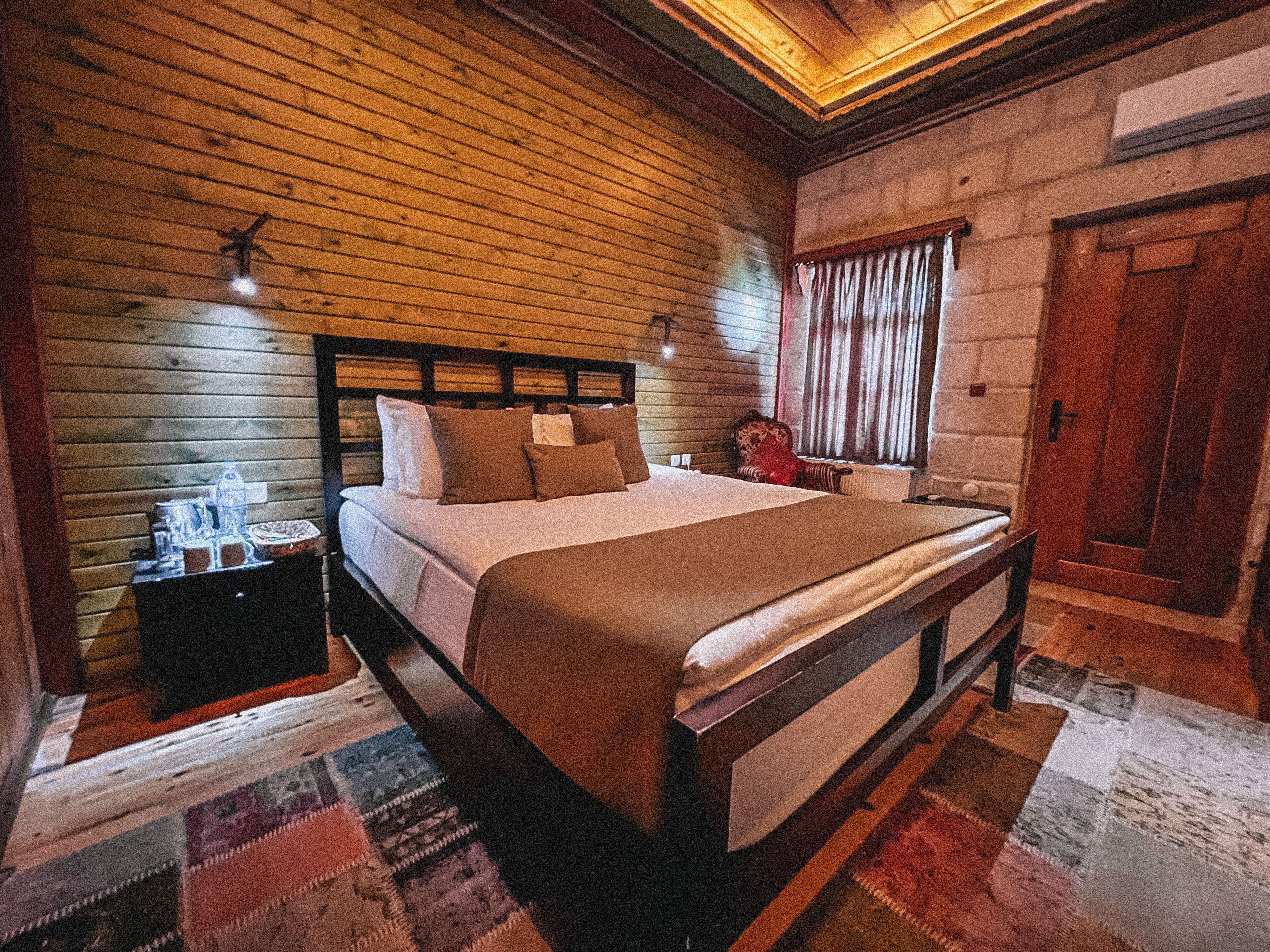 Quarto com painéis de madeira, cama, mesas de cabeceira e uma janela com cortinas.