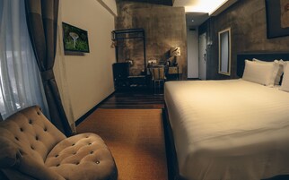 Quarto de hotel com uma cama grande, uma poltrona estofada, uma mesa de trabalho com cadeira e uma televisão na parede.