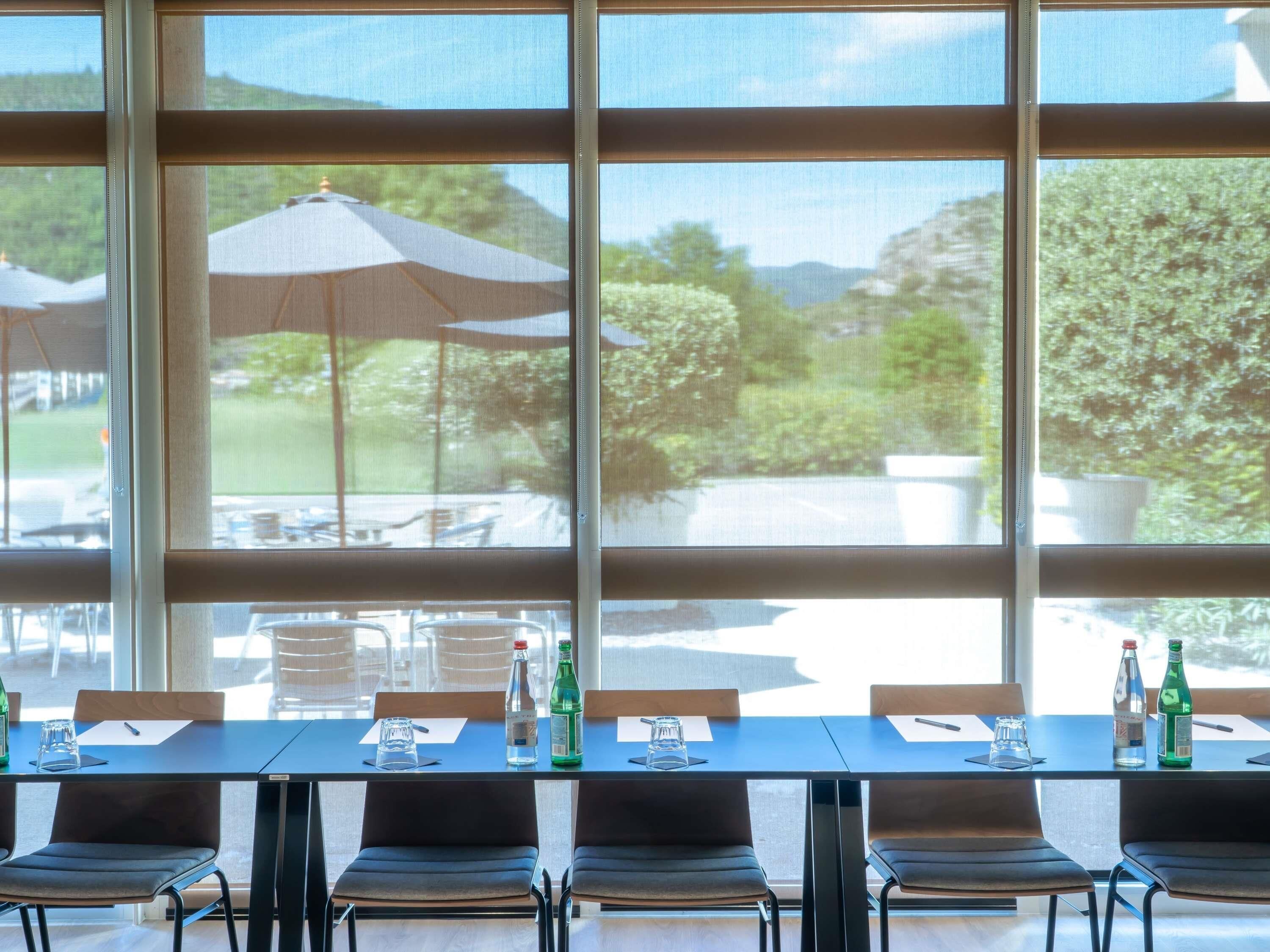 Sala de conferências com uma ampla janela que oferece vista para um pátio externo com guarda-sóis e mesas.