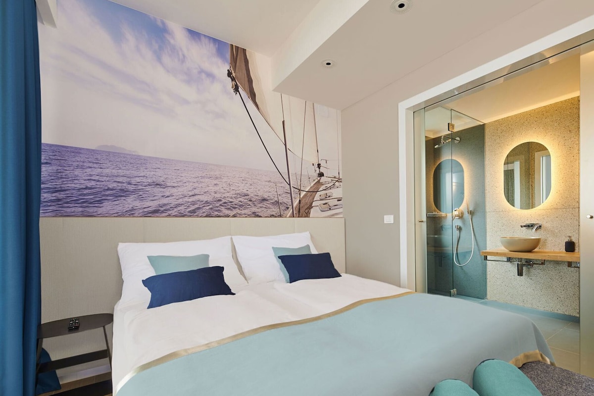 Quarto com cama, almofadas, painel decorativo na parede, banheiro com pia e espelho.