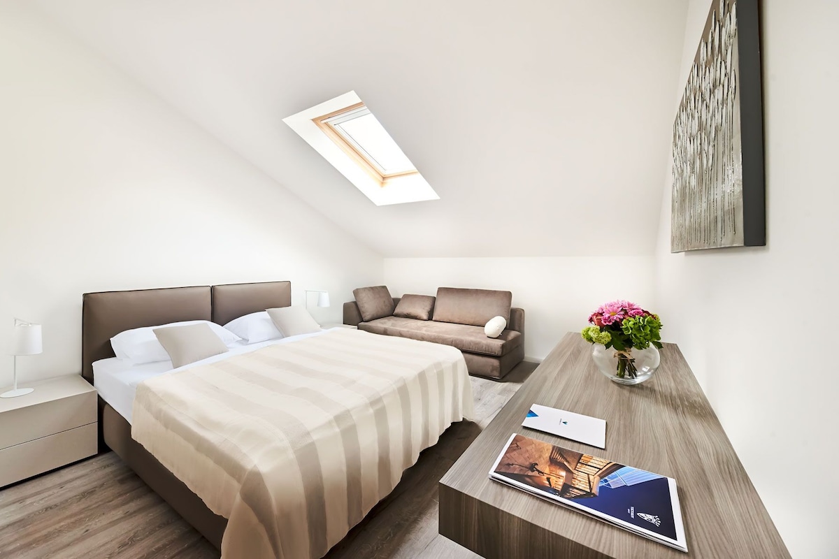 Quarto de hotel moderno com cama, sofá, escrivaninha com um vaso de flores e claraboia.