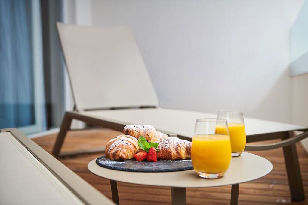Disposição para o pequeno-almoço com croissants, morangos e sumo de laranja sobre uma mesa num quarto com uma cadeira e uma janela.