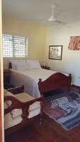 Quarto com cama de madeira, mesa de cabeceira, cadeira, ventilador de teto, janela com grades e um quadro na parede.