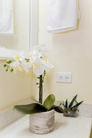 Um banheiro com pia branca, espelho e uma orquídea em vaso.