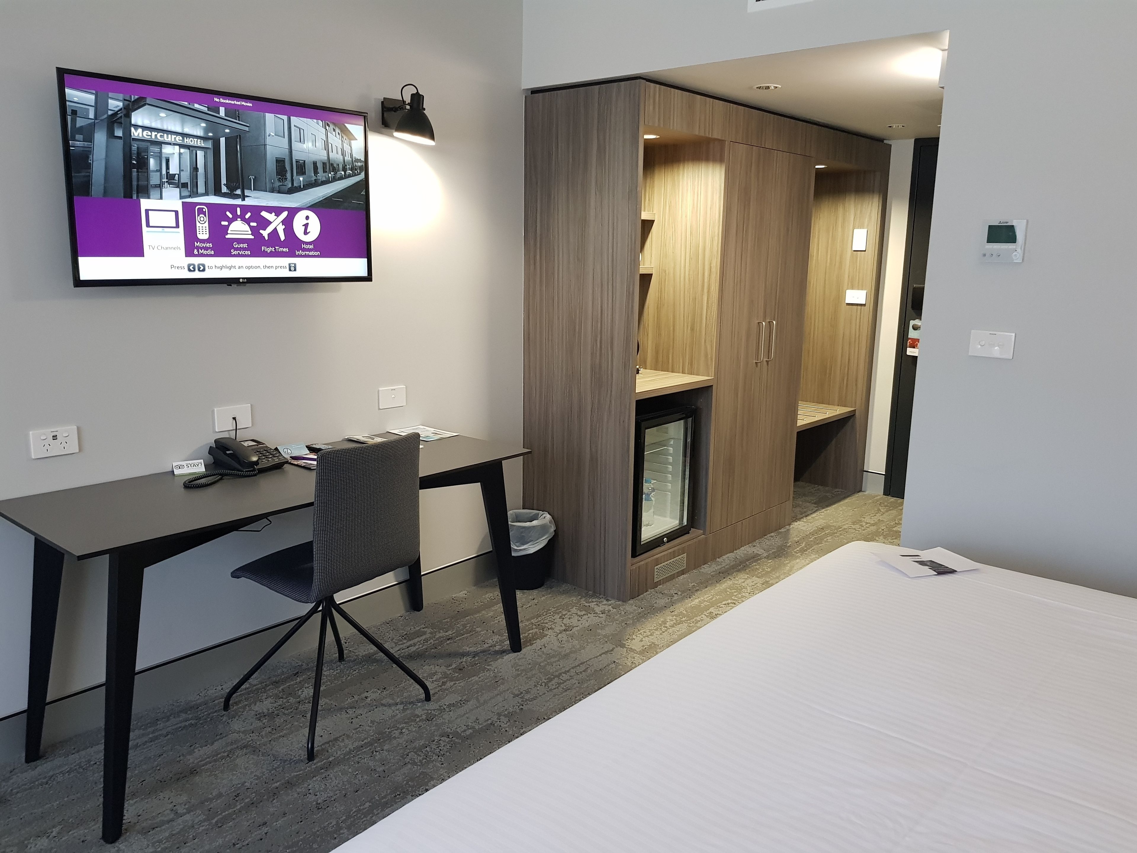 Quarto de hotel com escrivaninha, cadeira, televisão, guarda-roupa e cama.
