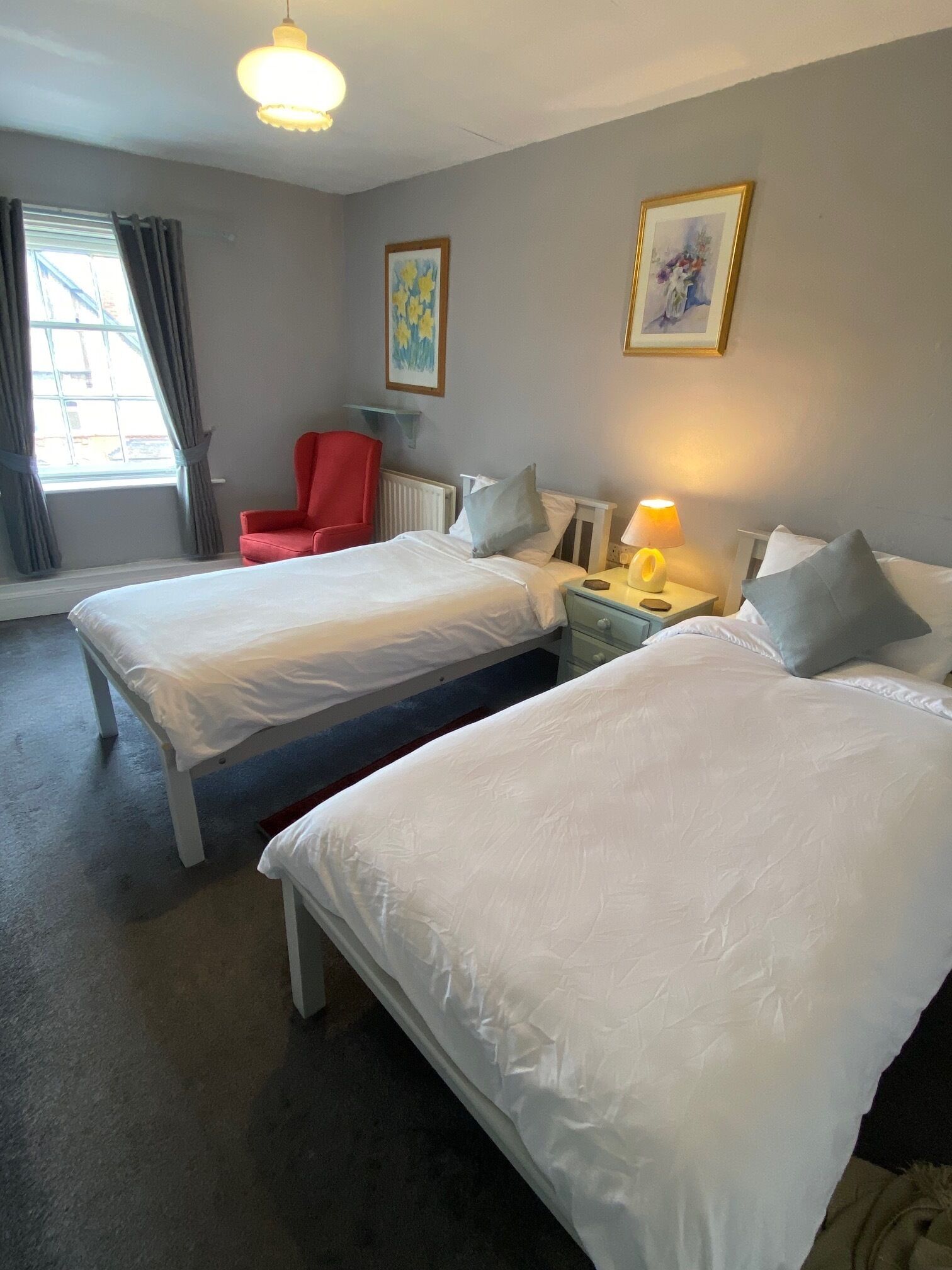 Quarto de hotel com duas camas, uma cadeira vermelha, uma mesinha de cabeceira com abajur e duas fotos emolduradas na parede.