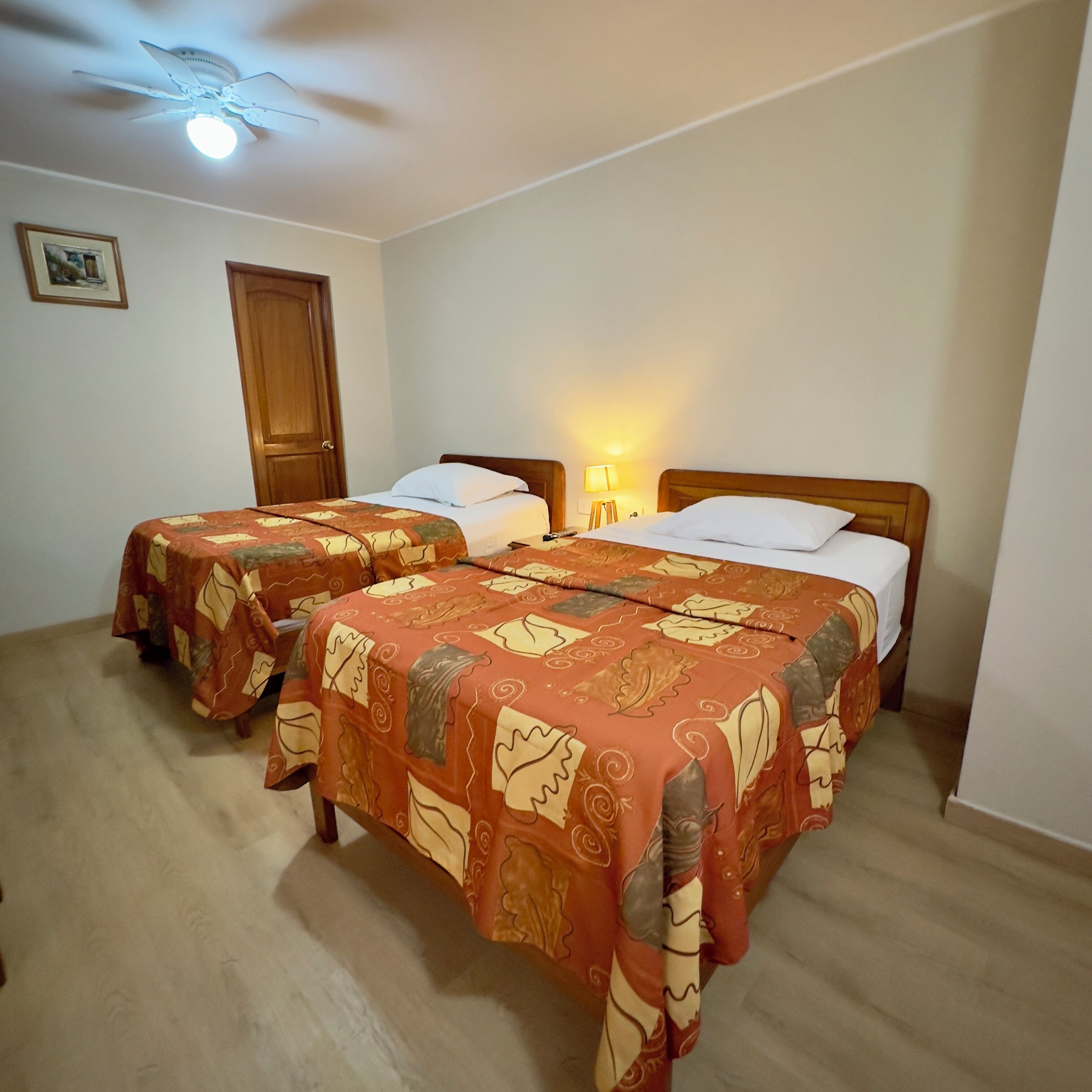 Quarto de hotel com duas camas, cada uma com colcha estampada, porta de madeira, ventilador de teto e quadro pendurado na parede.