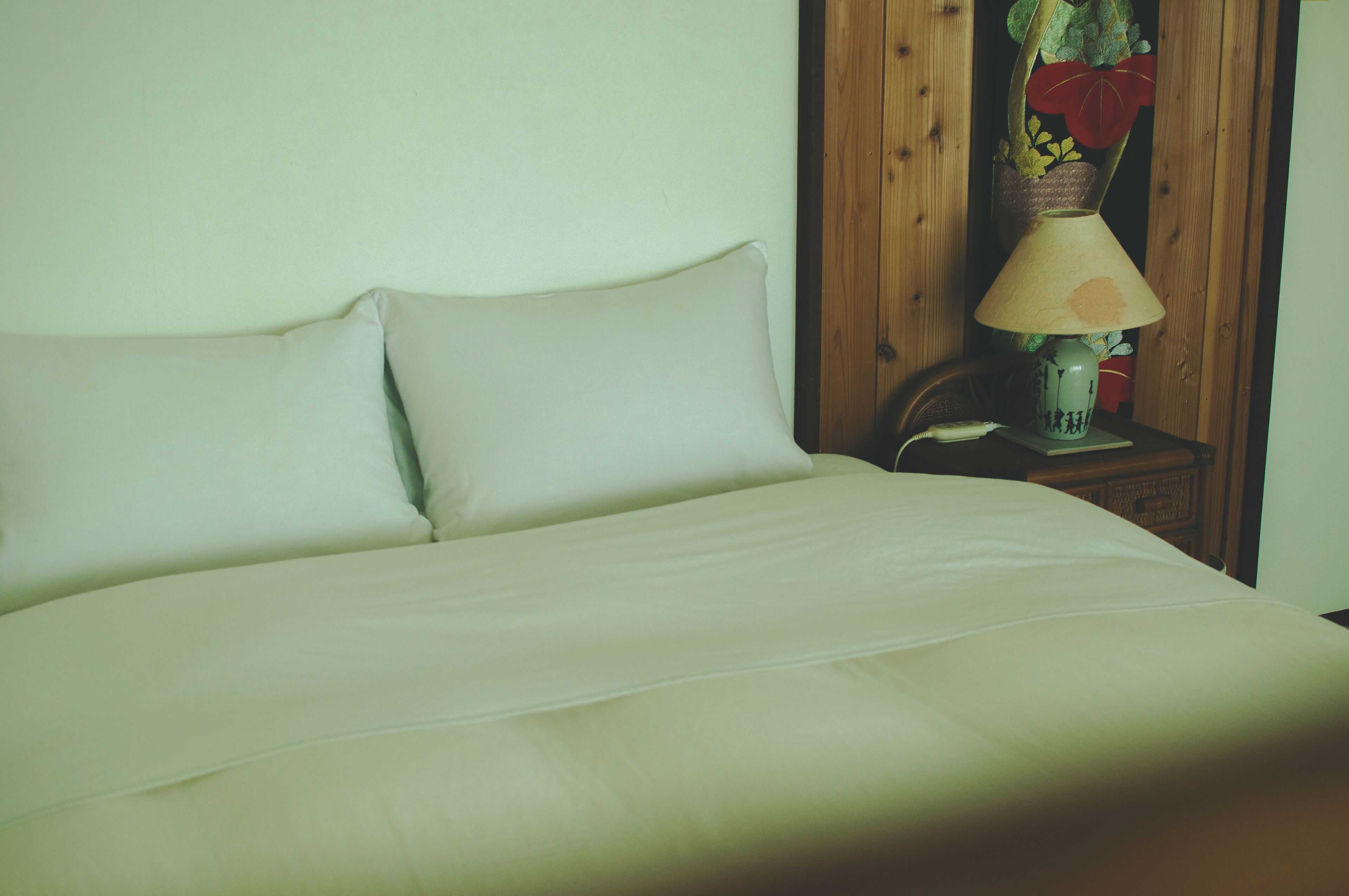 Uma cama com roupa de cama branca, uma mesa de cabeceira de madeira com uma luminária e uma parede com decoração floral.