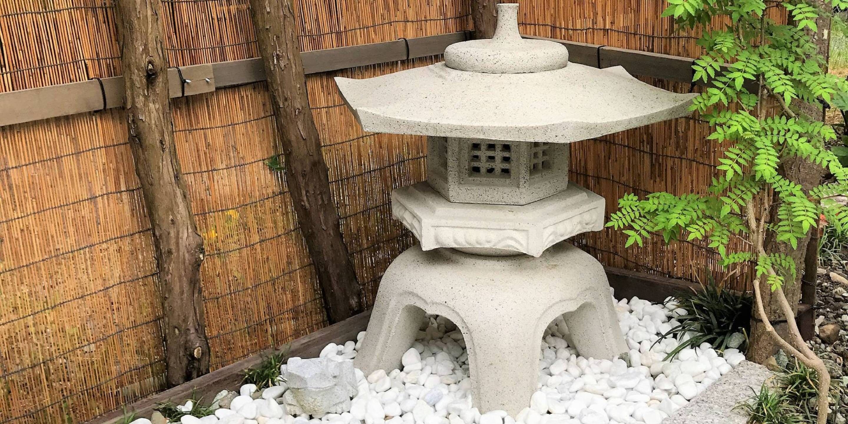 Um lanterne de pedra com design escalonado, cercado por pedrinhas brancas e vegetação, com um fundo de cerca de bambu.