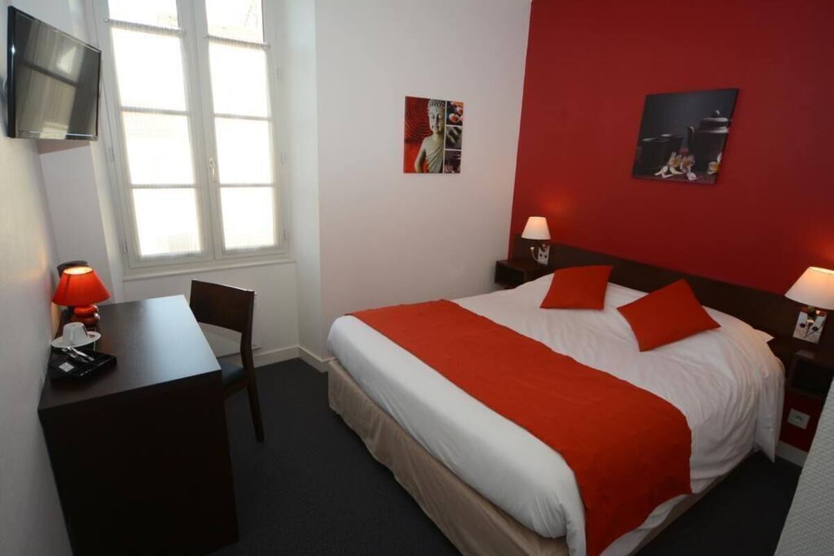 Quarto de hotel com uma cama, uma escrivaninha, uma cadeira, uma televisão e duas pinturas na parede.