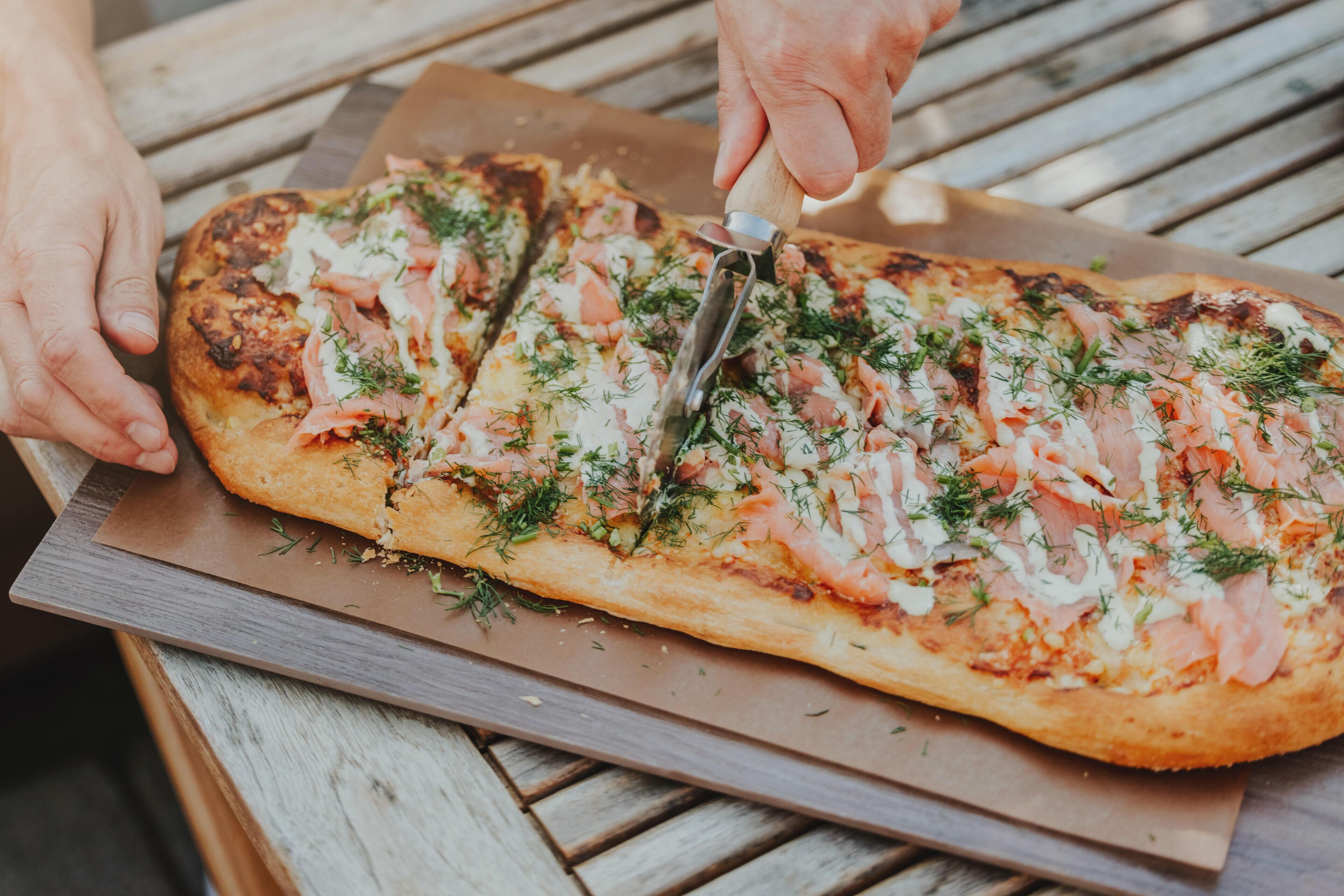 Uma pessoa cortando uma pizza longa e retangular sobre uma mesa de madeira.