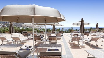 Área de lounge no terraço com espreguiçadeiras e guarda-sóis, com vista para a costa.