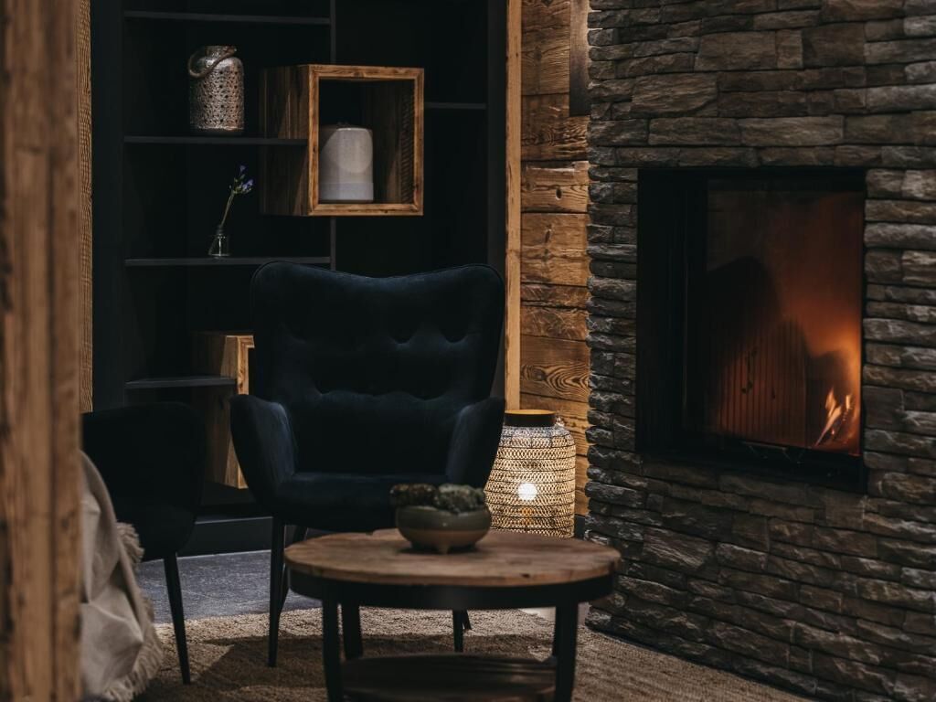 Um quarto acolhedor com lareira de pedra, uma poltrona azul escura, uma mesa redonda pequena e uma estante com objetos decorativos.