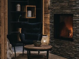 Um quarto acolhedor com lareira de pedra, uma poltrona azul escura, uma mesa redonda pequena e uma estante com objetos decorativos.