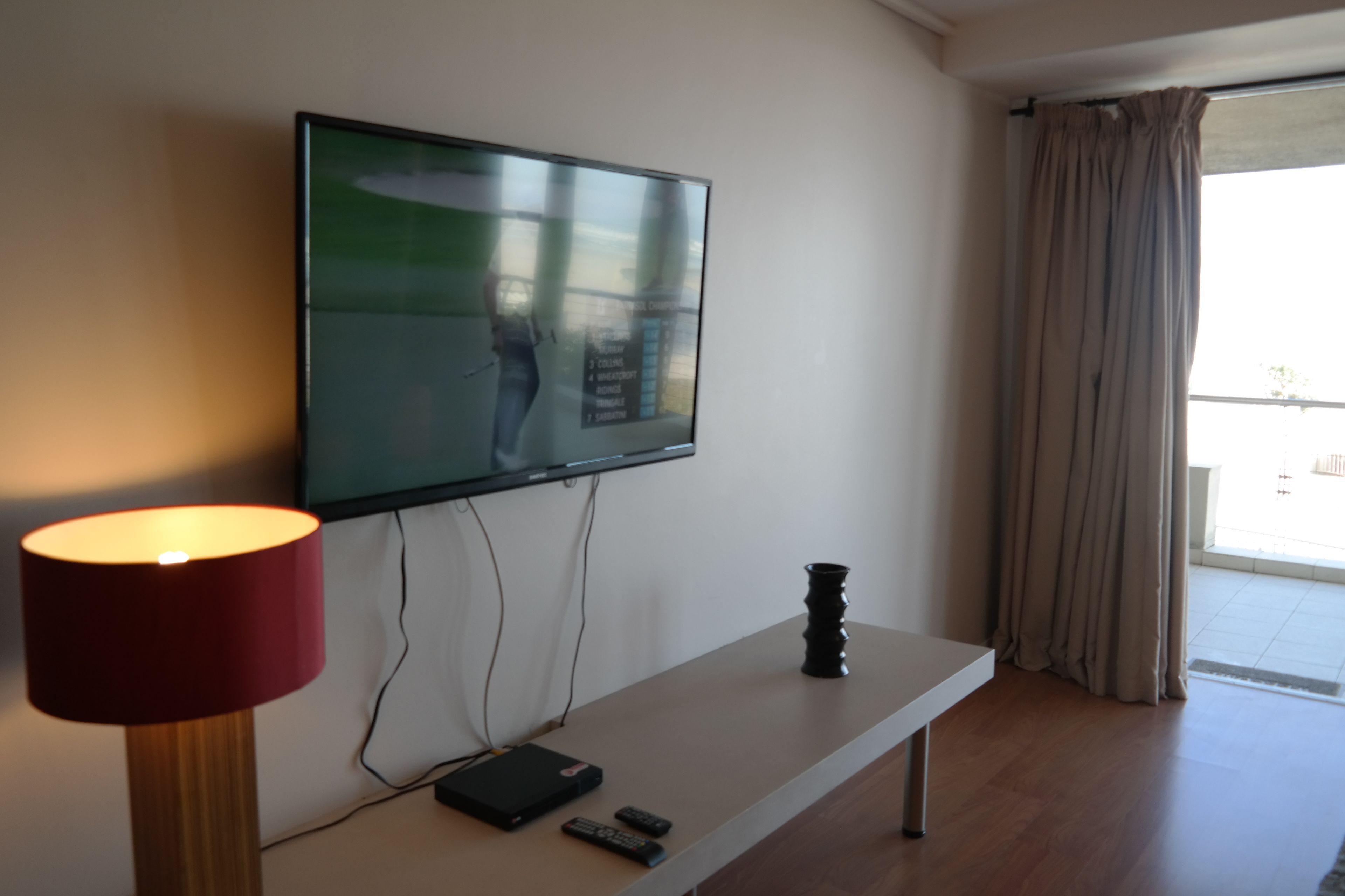 Quarto com televisão de tela plana na parede, luminária de chão, mesa pequena com controle remoto e janela com cortinas.
