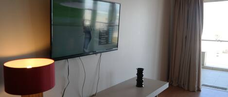 Quarto com televisão de tela plana na parede, luminária de chão, mesa pequena com controle remoto e janela com cortinas.