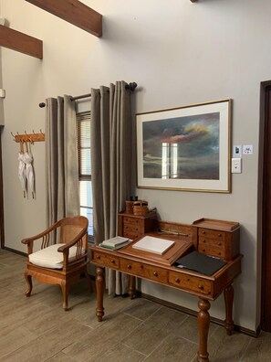 Um quarto com uma escrivaninha de madeira, uma cadeira, uma janela com cortinas, um cabideiro e um quadro na parede.