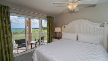 Quarto de hotel com cama, ventilador de teto, mesa de cabeceira com abajur, uma cadeira e vista para o oceano através de grandes janelas.