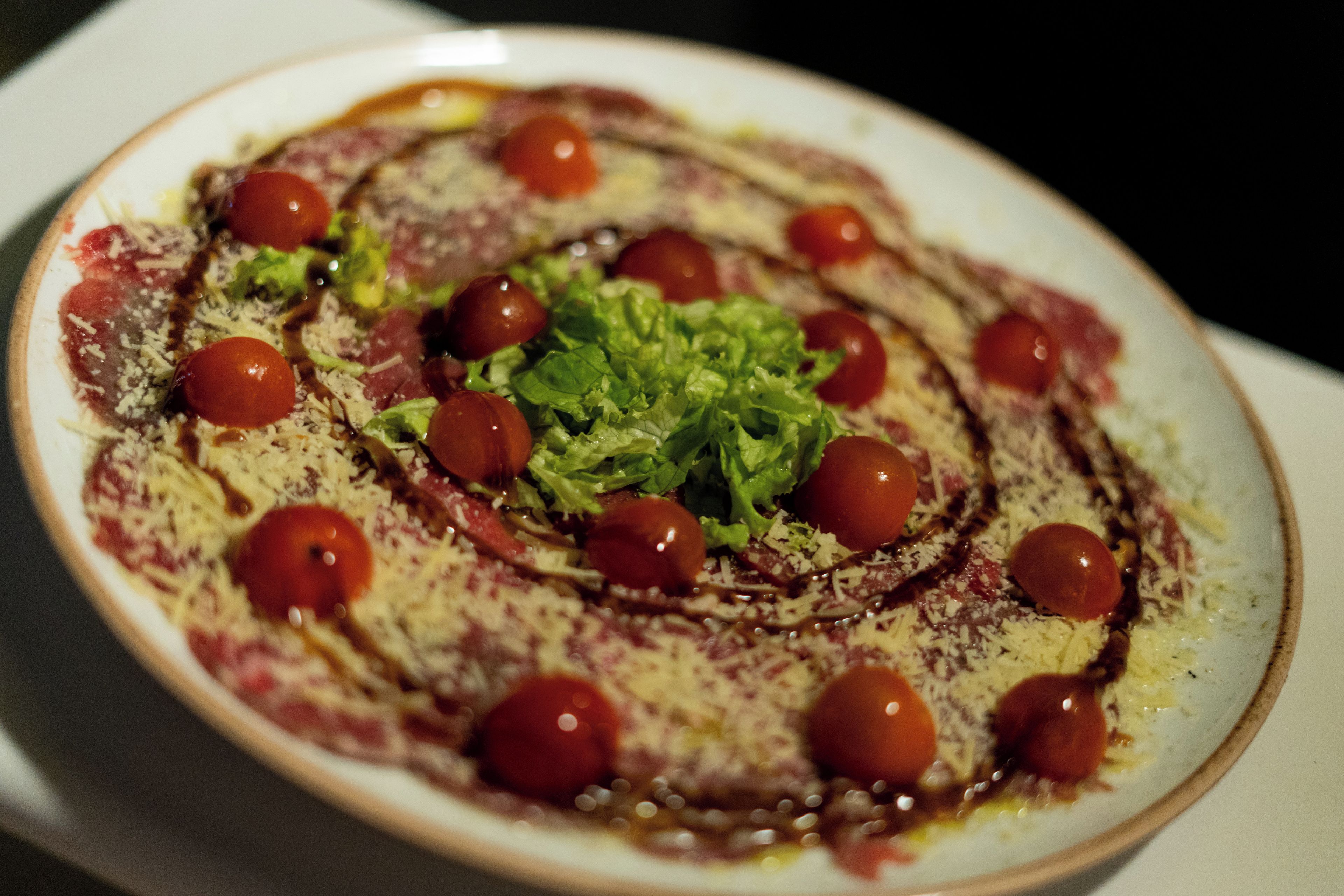 Prato de comida com guarnição central de folhas verdes, rodeado por tomates vermelhos igualmente espaçados e um fio de molho.